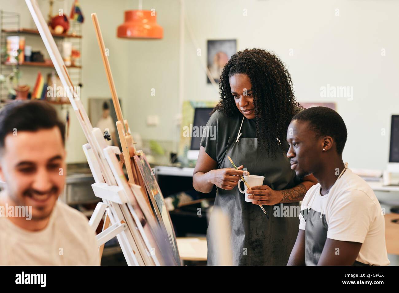 Female tutor looking at painting on easel by male student sitting in ...