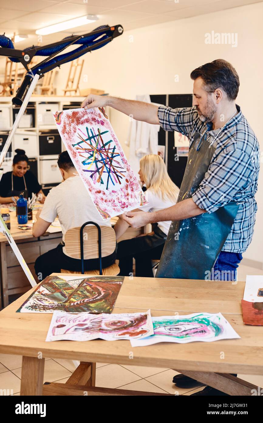 Mature male instructor examining painting while students practicing in ...