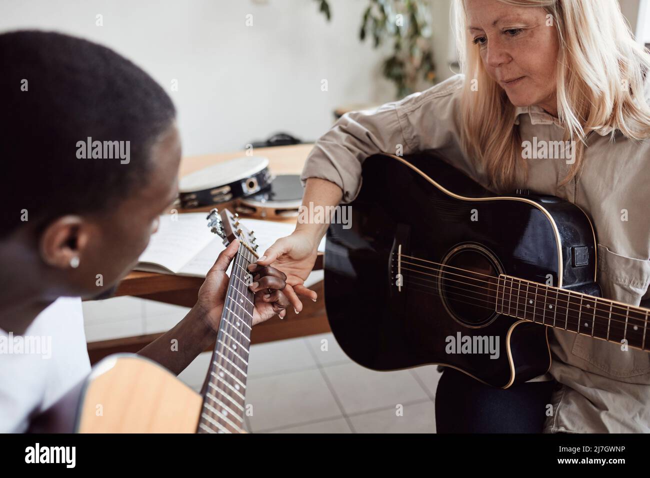 Blond female tutor looking at male student playing guitar in classroom ...