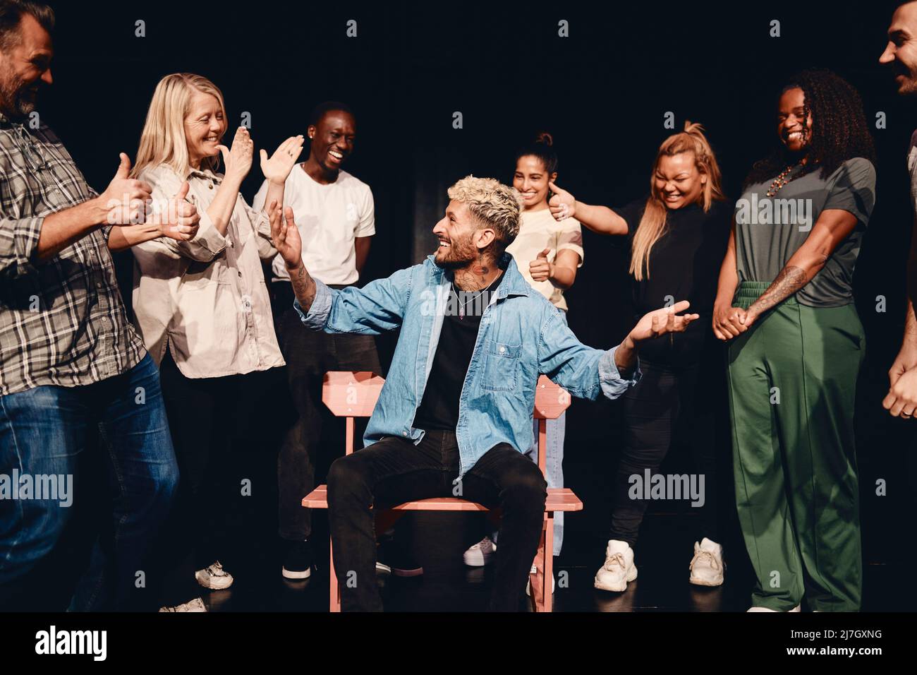 Happy young man sitting on chair amidst multiracial stage performers in ...