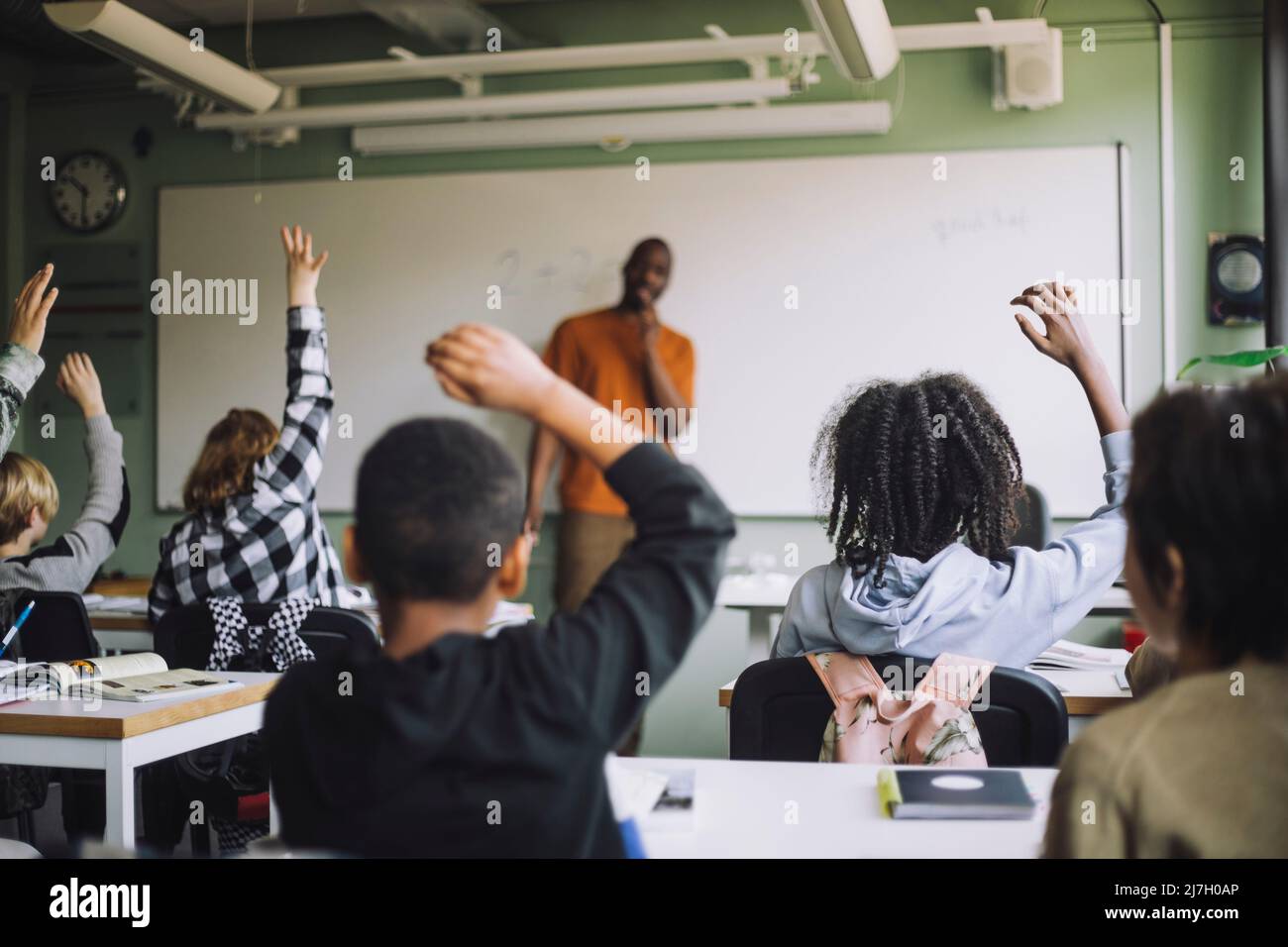 Rear view of multiracial students raising hands in classroom during ... Rear view of multiracial students raising hands in classroom during ...