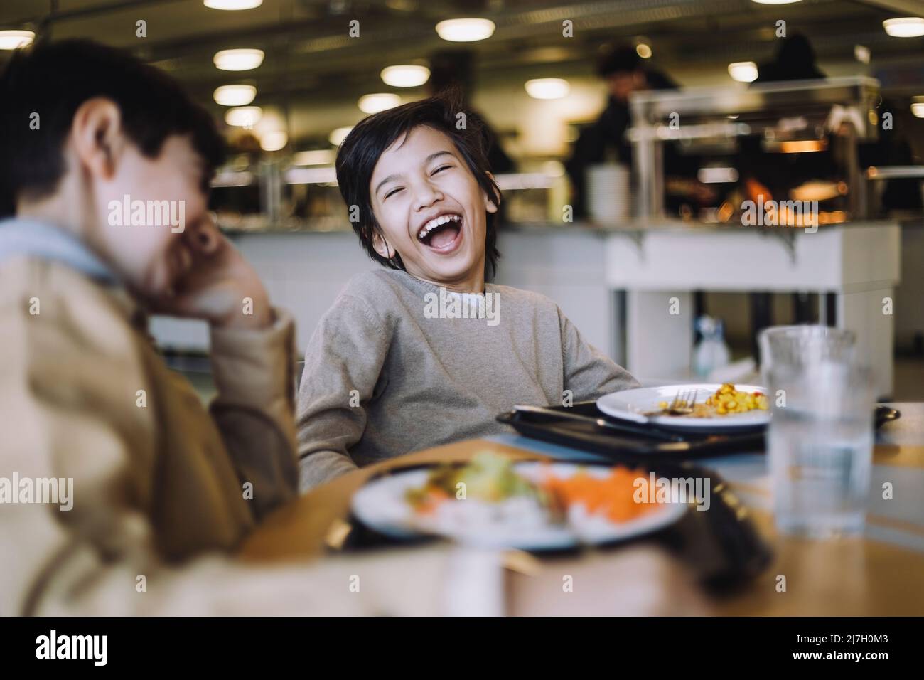 Boy laughing while sitting with friend during lunch break at school ...