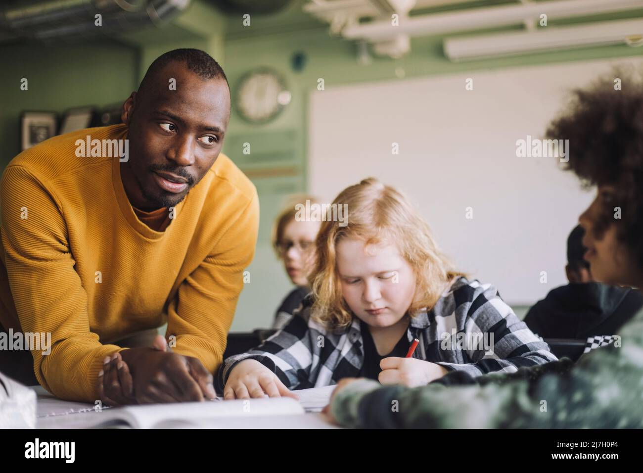 Serious teacher looking at student while leaning at desk in classroom ...