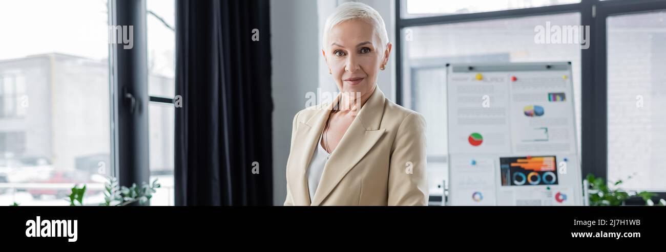 senior banker smiling at camera near flip chart with graphs on blurred ...
