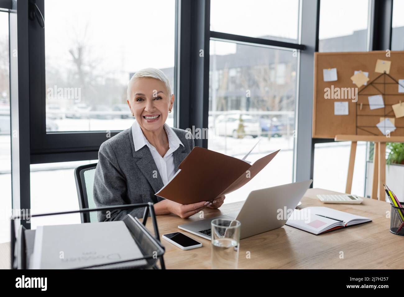 happy banker with folder smiling at camera near laptop and cellphone ...