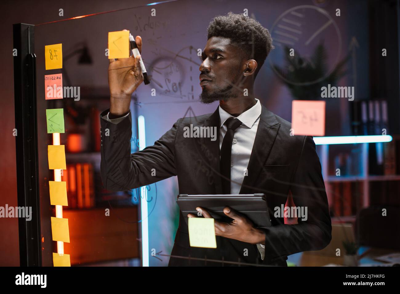 Focused African male CEO writes on the glass office board with sticky ...
