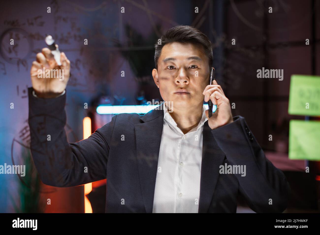 Focused Asian male ceo manager in formal wear writing on glass board ...