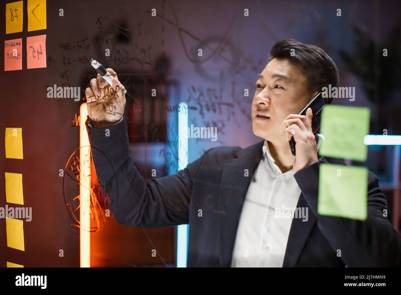 Focused Asian male ceo manager in formal wear writing on glass board ...