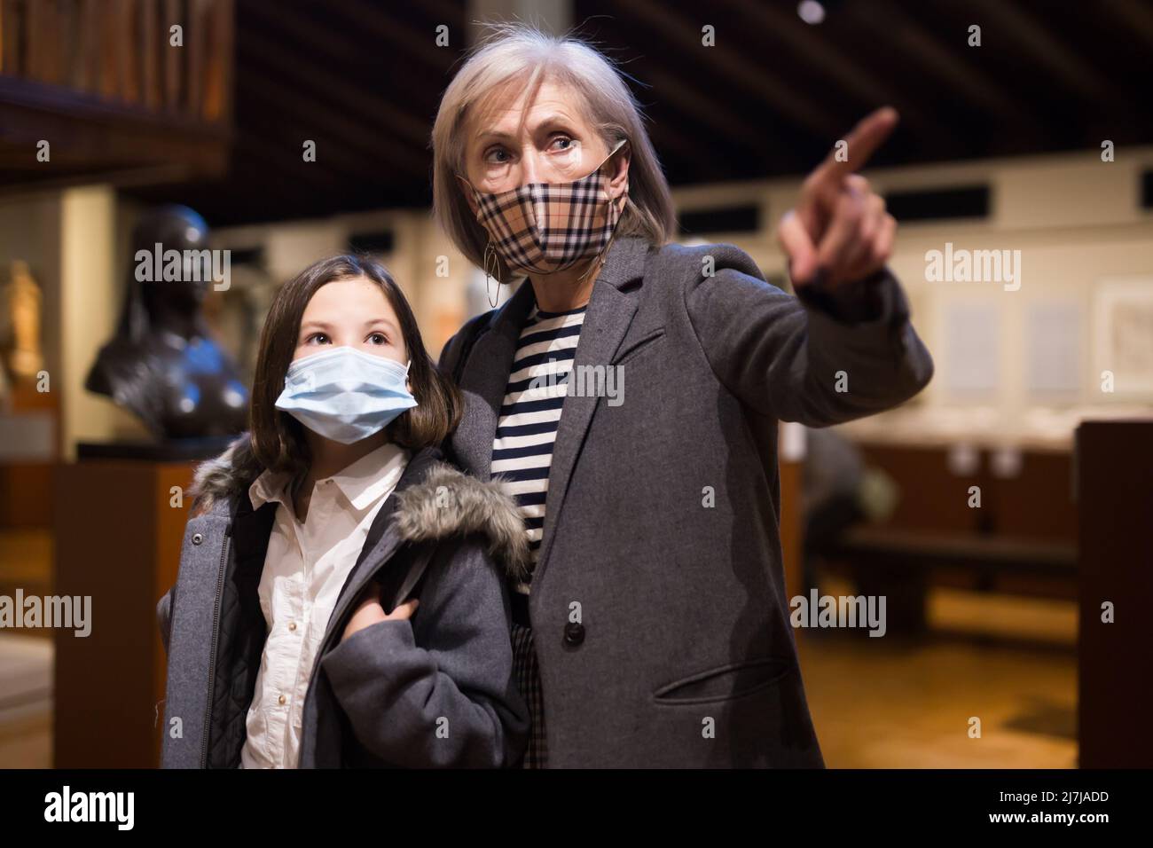 Preteen girl and senior woman in protective masks exploring artworks in ...