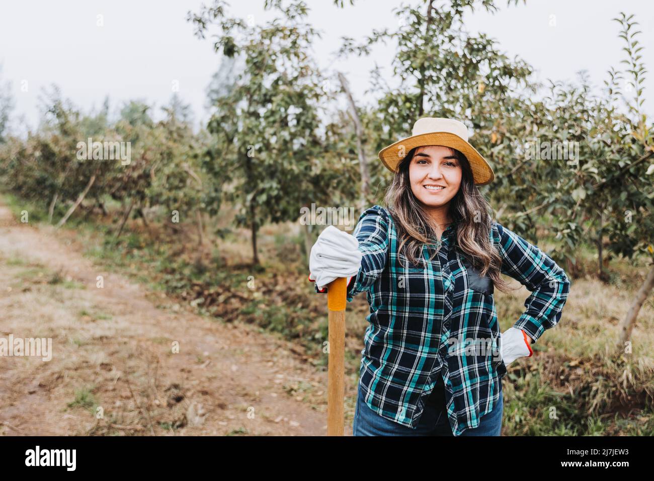 Young smiling latin peasant woman using gardening gloves and leaning on ...