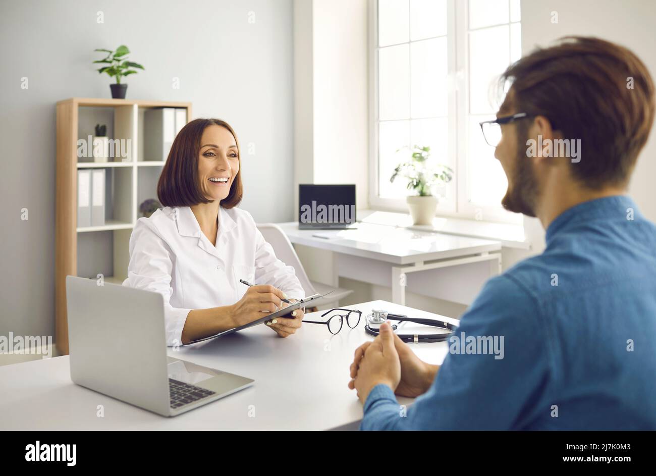 Happy doctor smiling and talking to patient sitting at desk in clinic ...