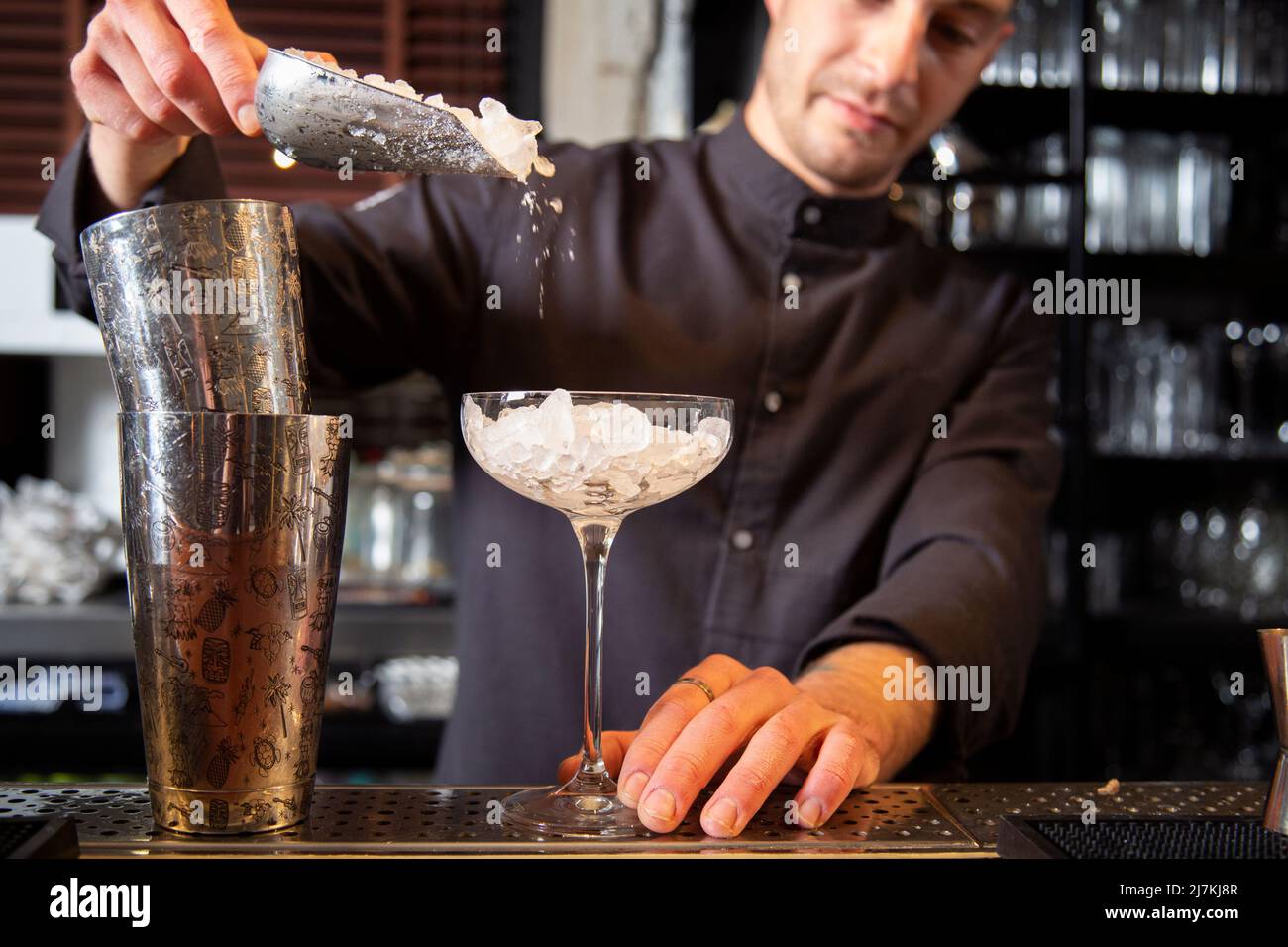 Crop young male barkeeper in black uniform pouring crushed ice from ...