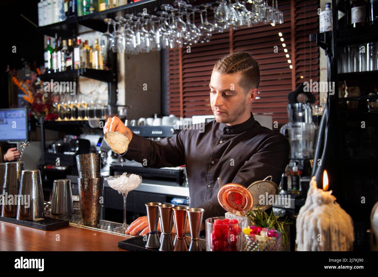 Concentrated young male barkeeper in black uniform pouring crushed ice ...