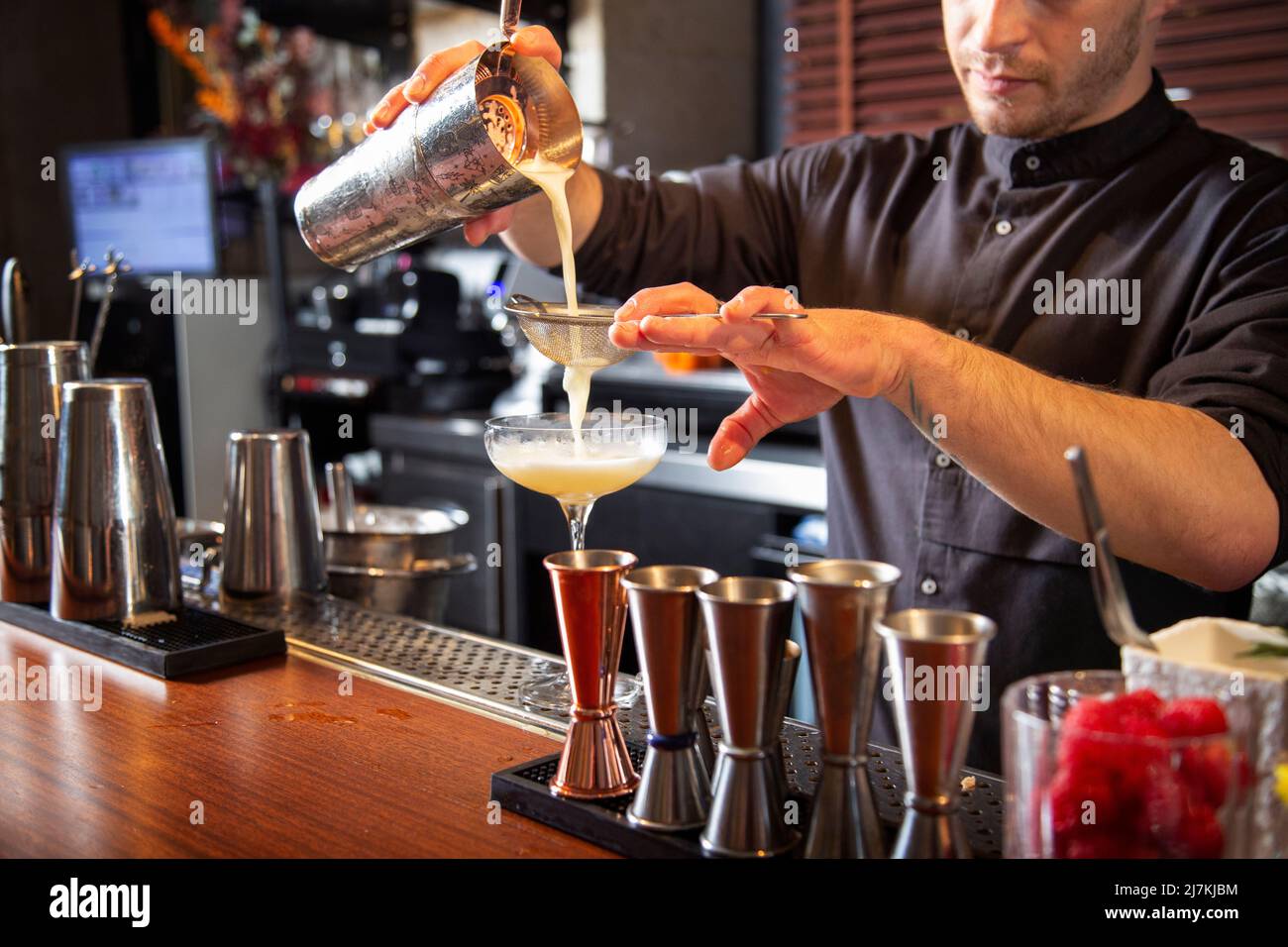 Crop bearded young male bartender in uniform pouring mixed cocktail ...