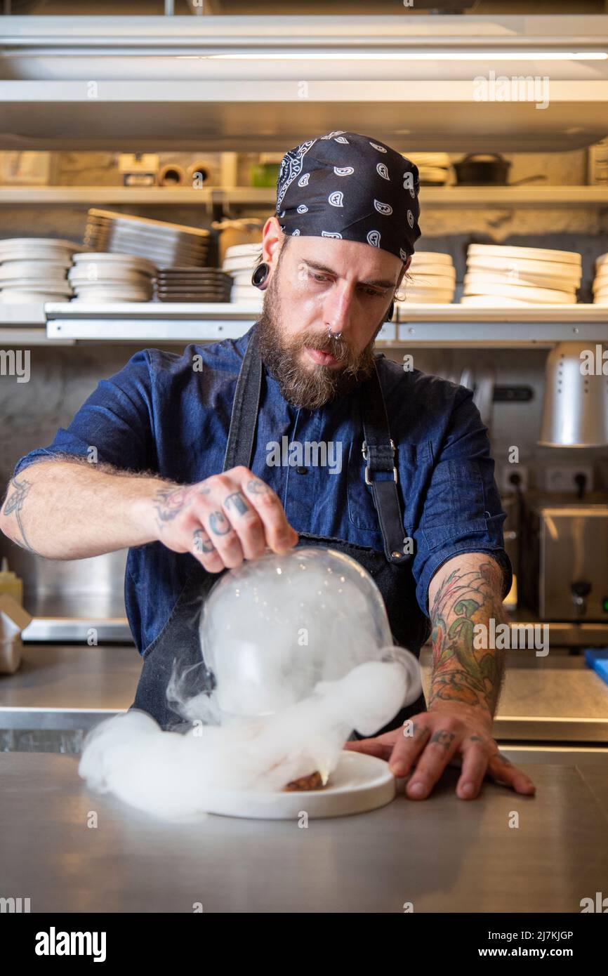 Serious focused bearded male chef looking at camera while using smoking ...