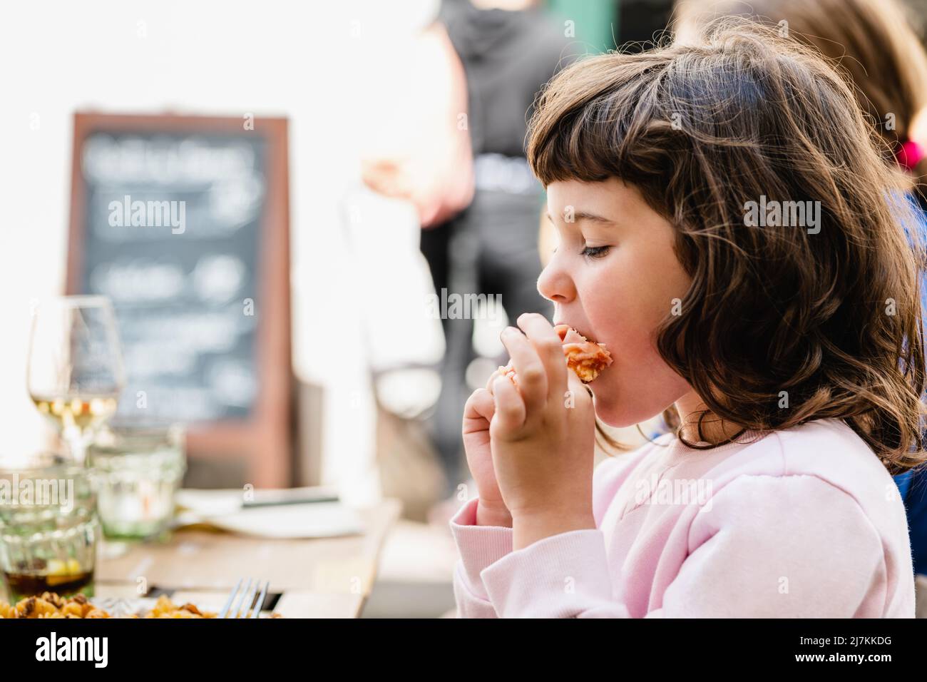 Side view of little girl with dark hair biting delicious pizza while ...