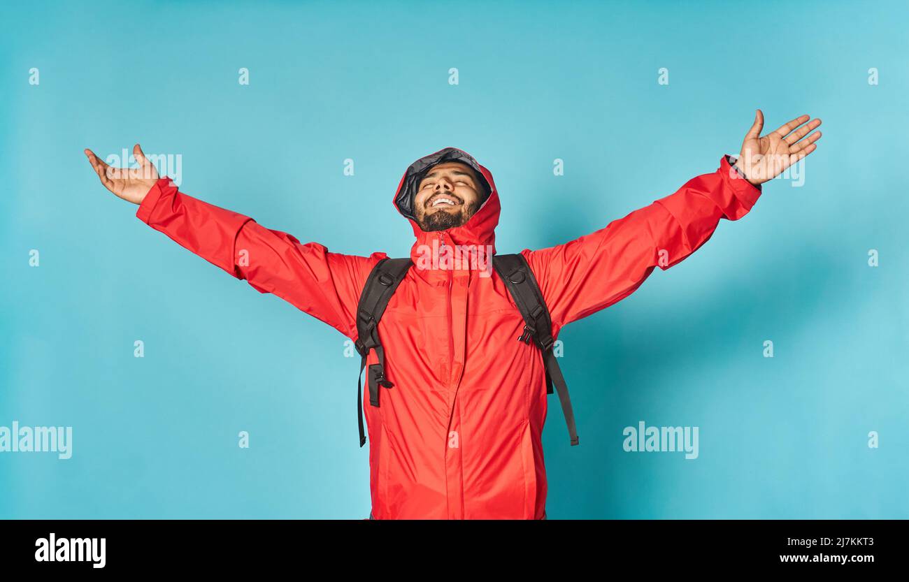 Happy bearded male tourist in red jacket and backpack with raised arms ...