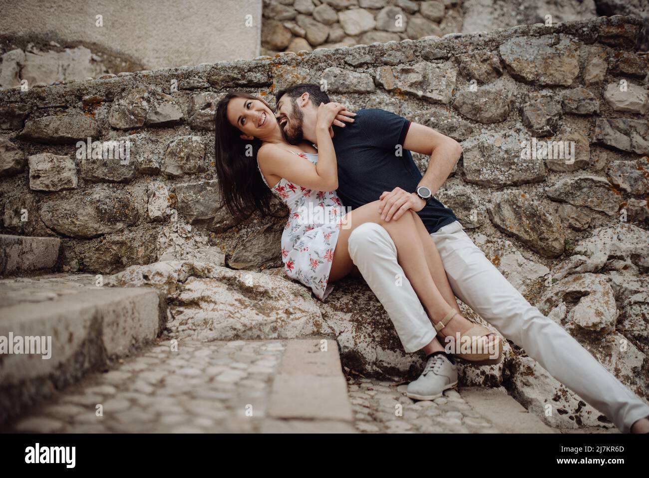Playful casual couple cuddling and sitting on cobbled city street on ...