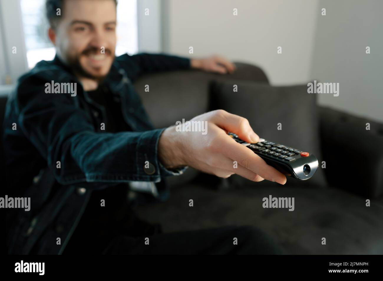 Happy young man wearing denim clothes with remote control switching channels on TV at home ...
