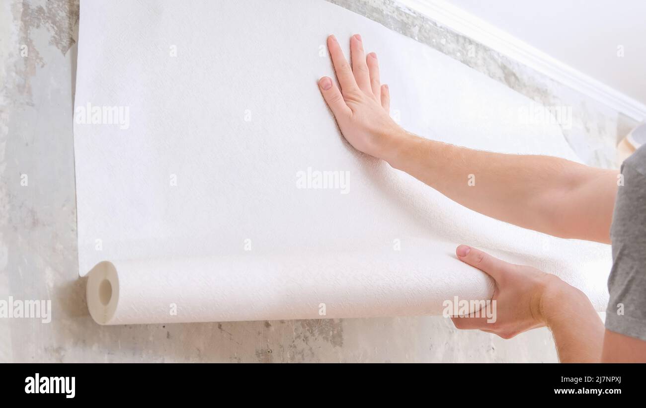 A man rolls out a roll of white wallpaper on the wall with glue ...