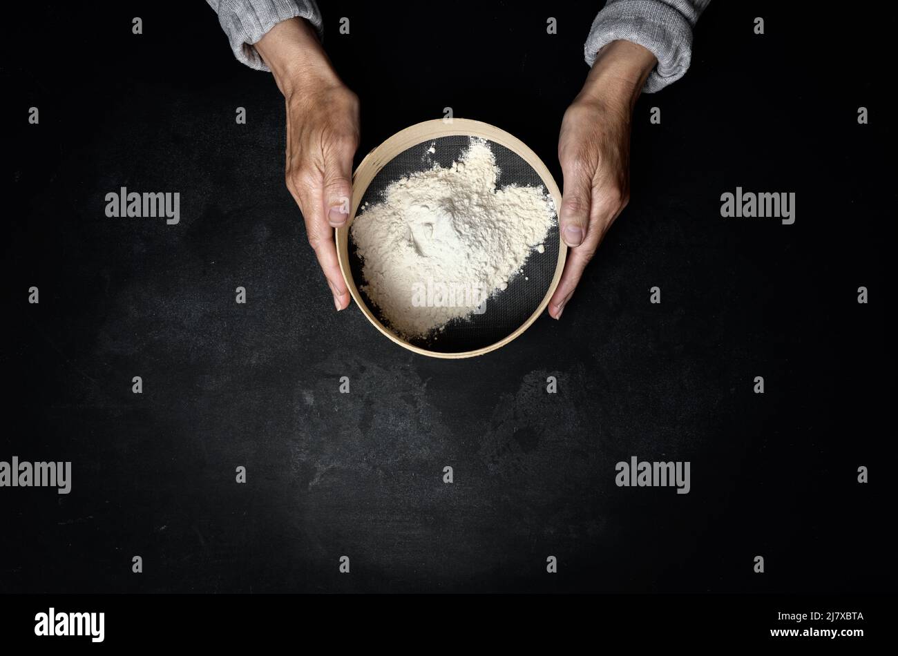 two female hands hold a round wooden sieve with wheat flour on a black ...