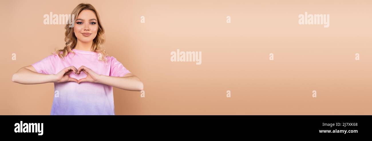 young woman smiling at camera and showing heart sign isolated on beige ...
