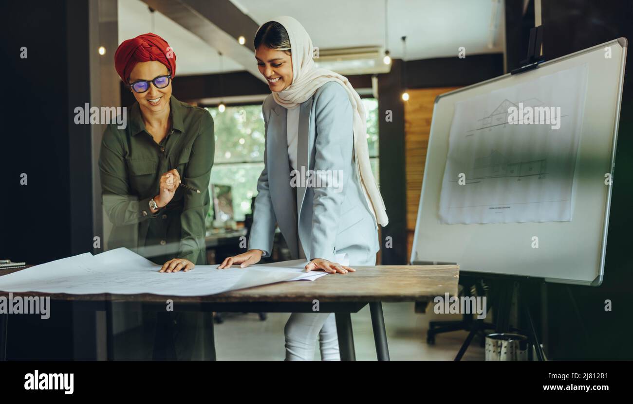 Cheerful female architects working on blueprint drawings in a modern office. Two Muslim ...