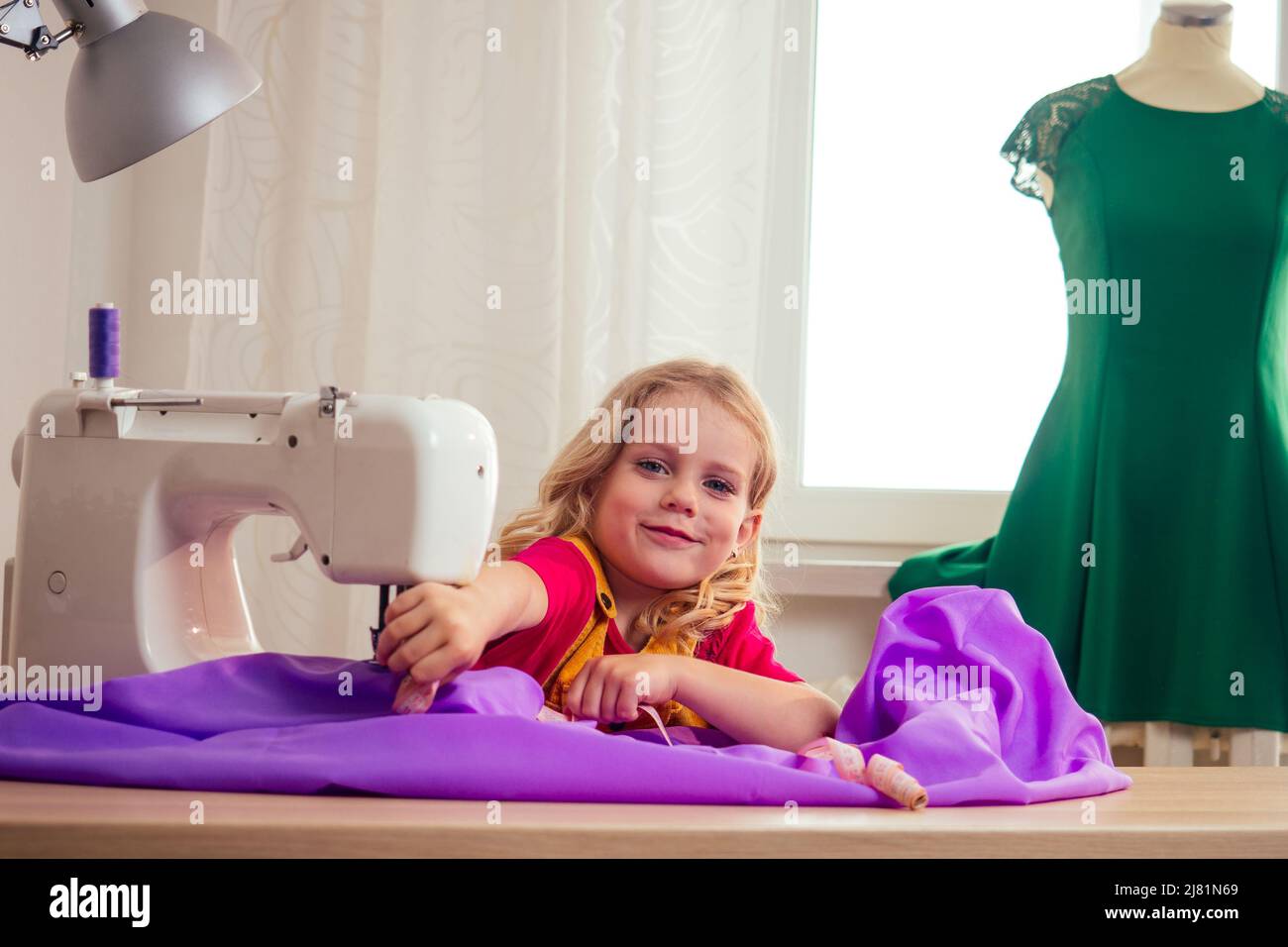 smiling little blonde girl at the table with sewing machine with ...