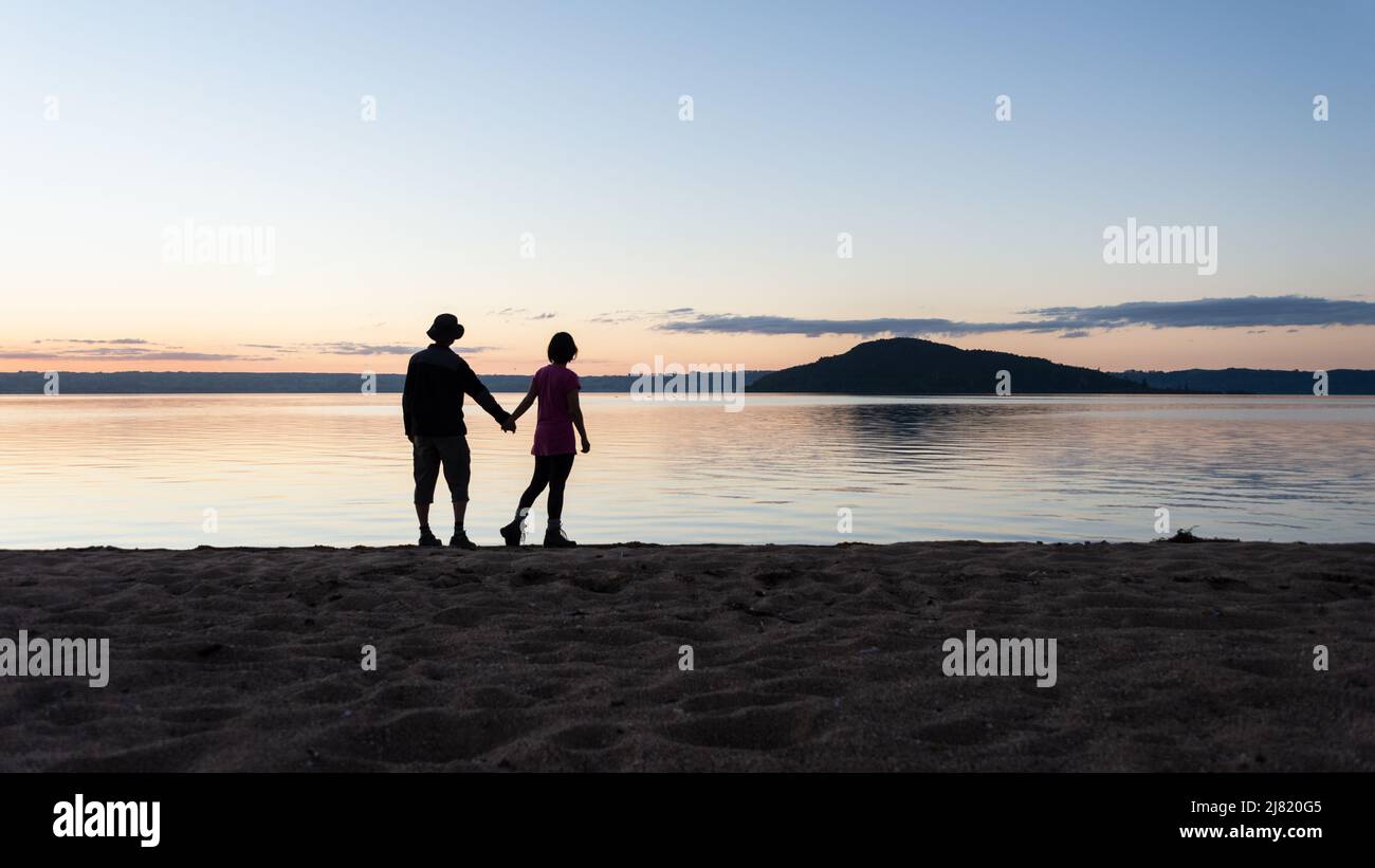 Two people walking by lake Rotorua at sunset, Mokoia Island in the ...