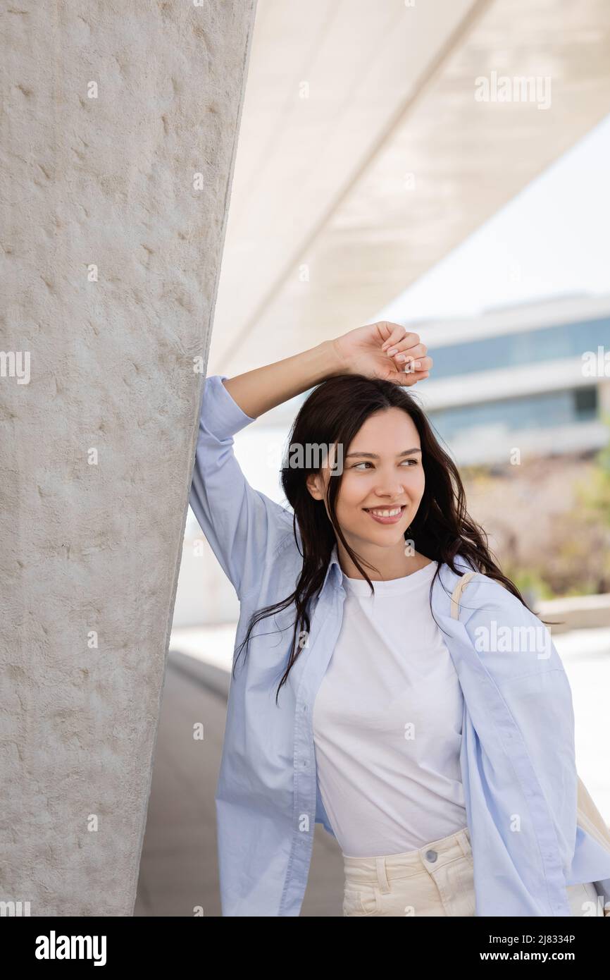 happy woman in blue shirt standing with hand above head near grey wall ...