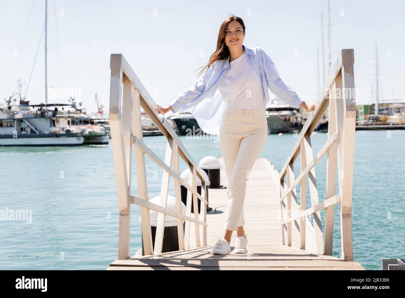 full length of smiling woman in white pants standing on pier in seaport ...