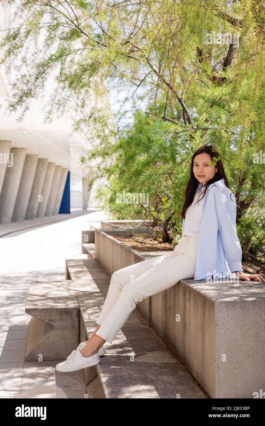 full length view of brunette woman in white pants sitting under trees ...