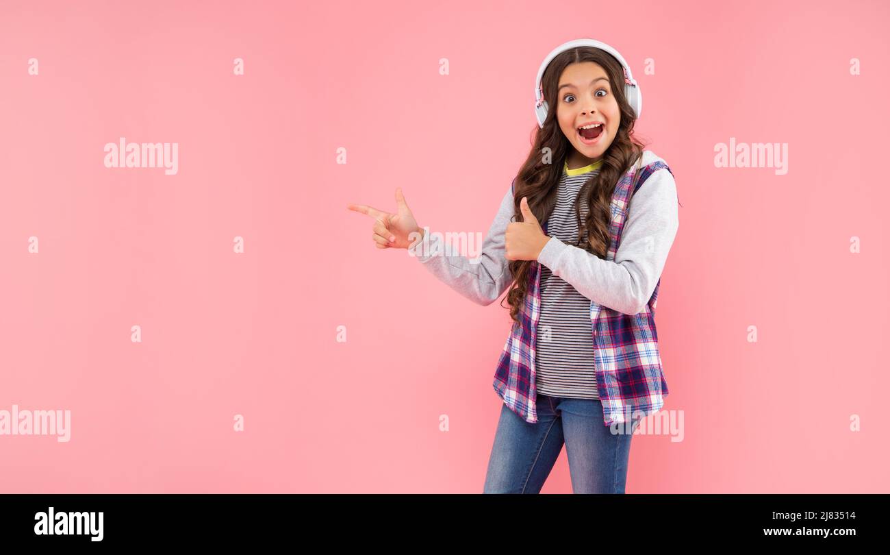 amazed kid listening music in earphones point finger on pink background ...
