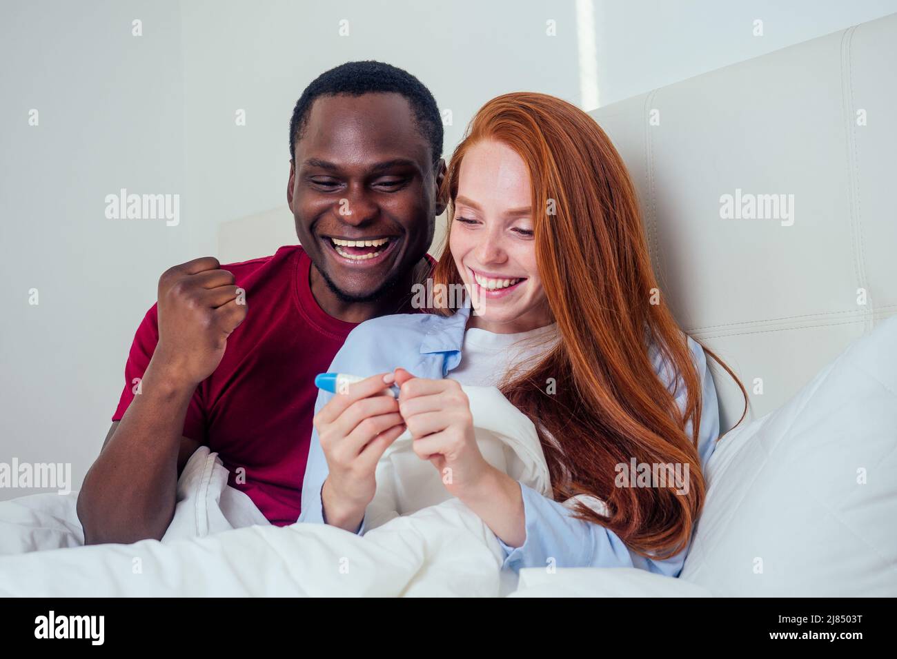 mixed race couple watching pregnancy test in bed. redhaired ginger ...