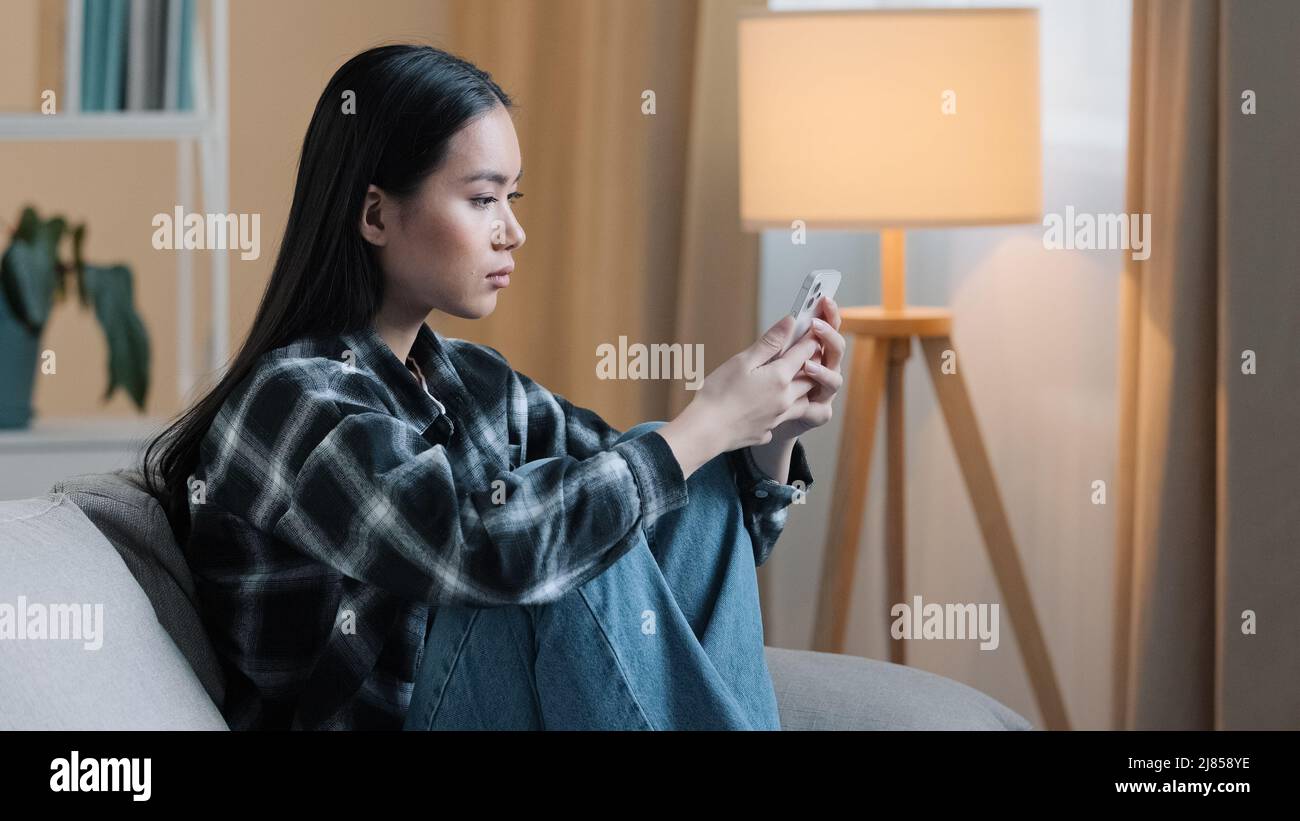 Side view of sad bored Asian woman girl sitting on sofa at home with ...