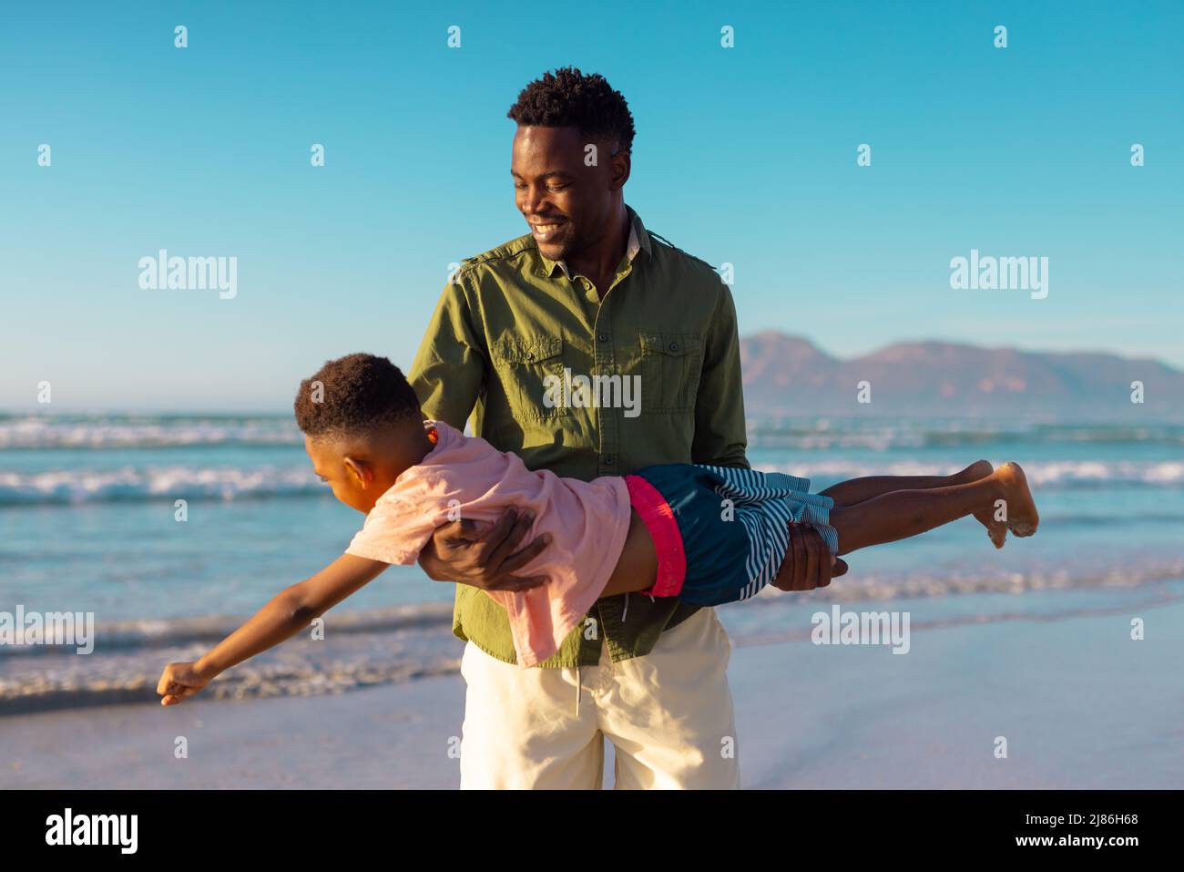 Smiling african american young man carrying playful son against sea and ...