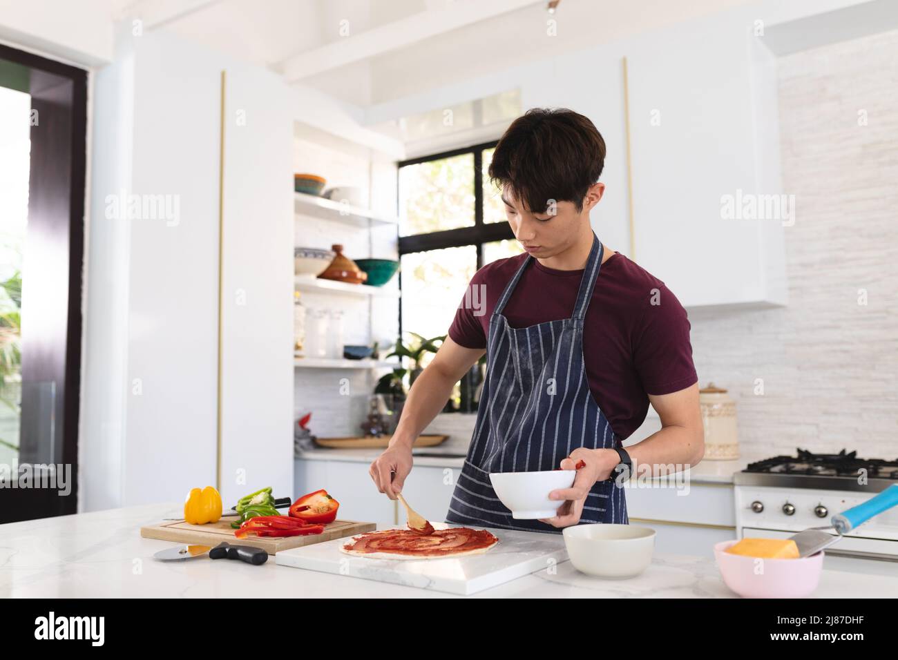 Asian teenage boy wearing apron spreading sauce on pizza while standing ...