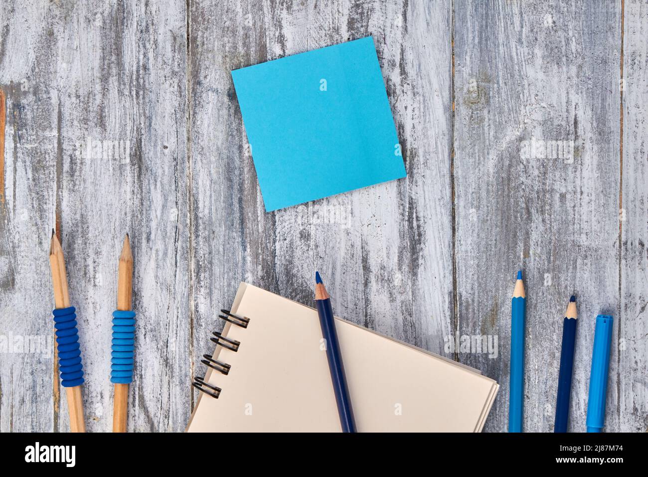Blank blue sticky note with pencils and notepad. Gray wood background ...