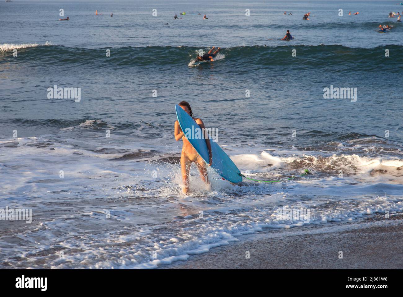 A blonde Caucasian woman exiting the ocean carrying a blue long surf ...