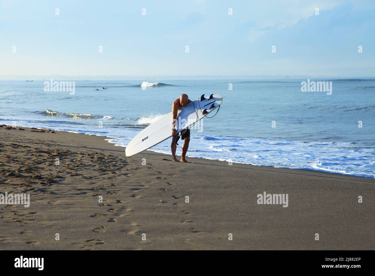 An elderly male Caucasian man carrying a longboard at Batu Bolong Beach ...