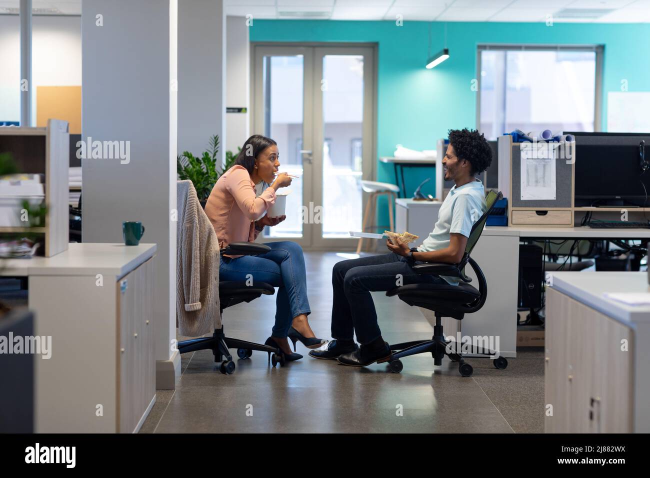 Multiracial professionals eating lunch together while sitting on chairs ...