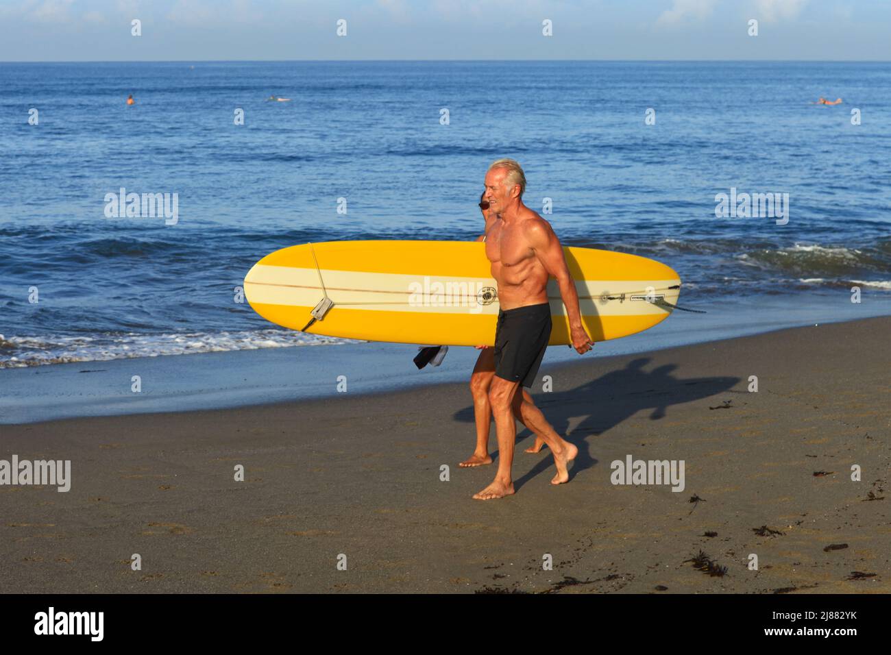 An elderly male Caucasian man carrying a yellow longboard at Batu ...