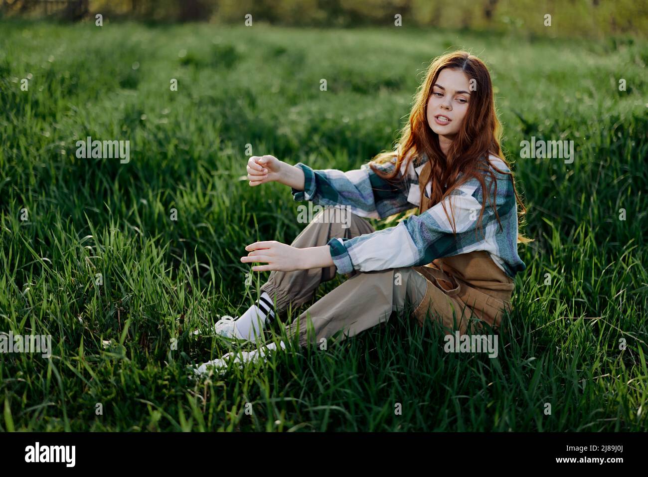 A young beautiful woman sits on the green grass in the park and looks ...