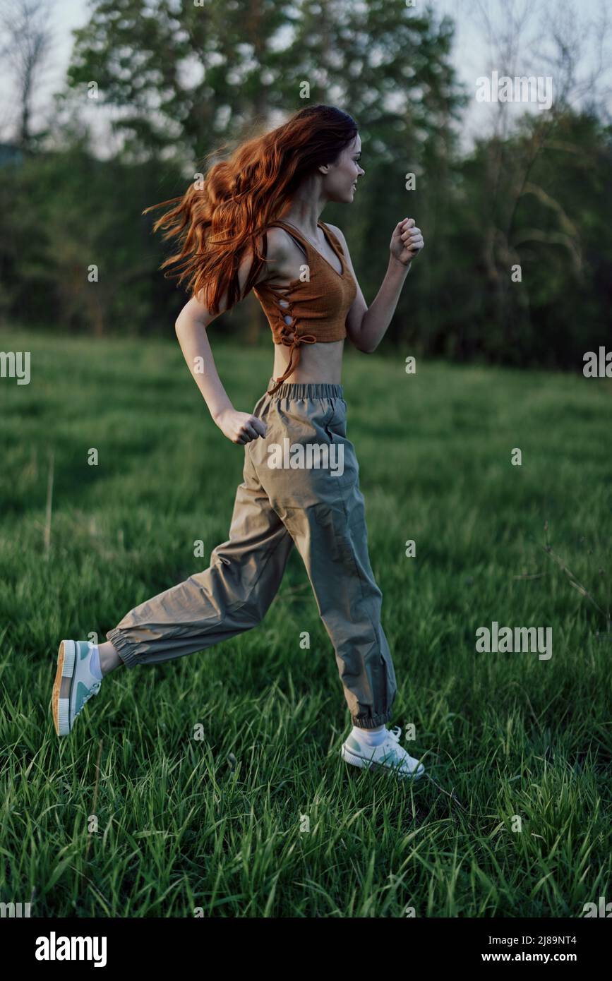 A woman with long red hair works out and runs on the green grass in the ...