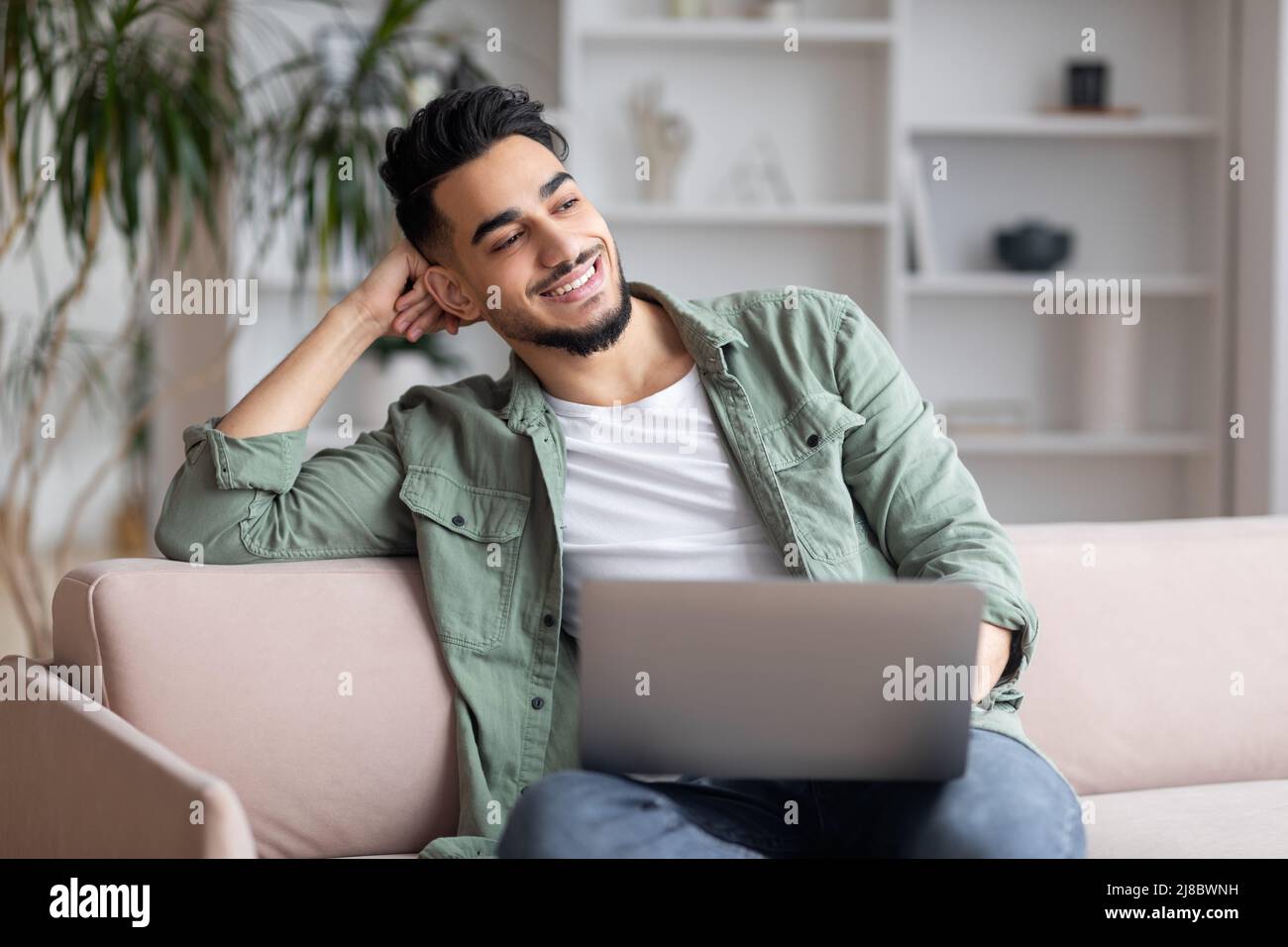 Smiling young muslim man with beard watch at free space, sits on sofa ...