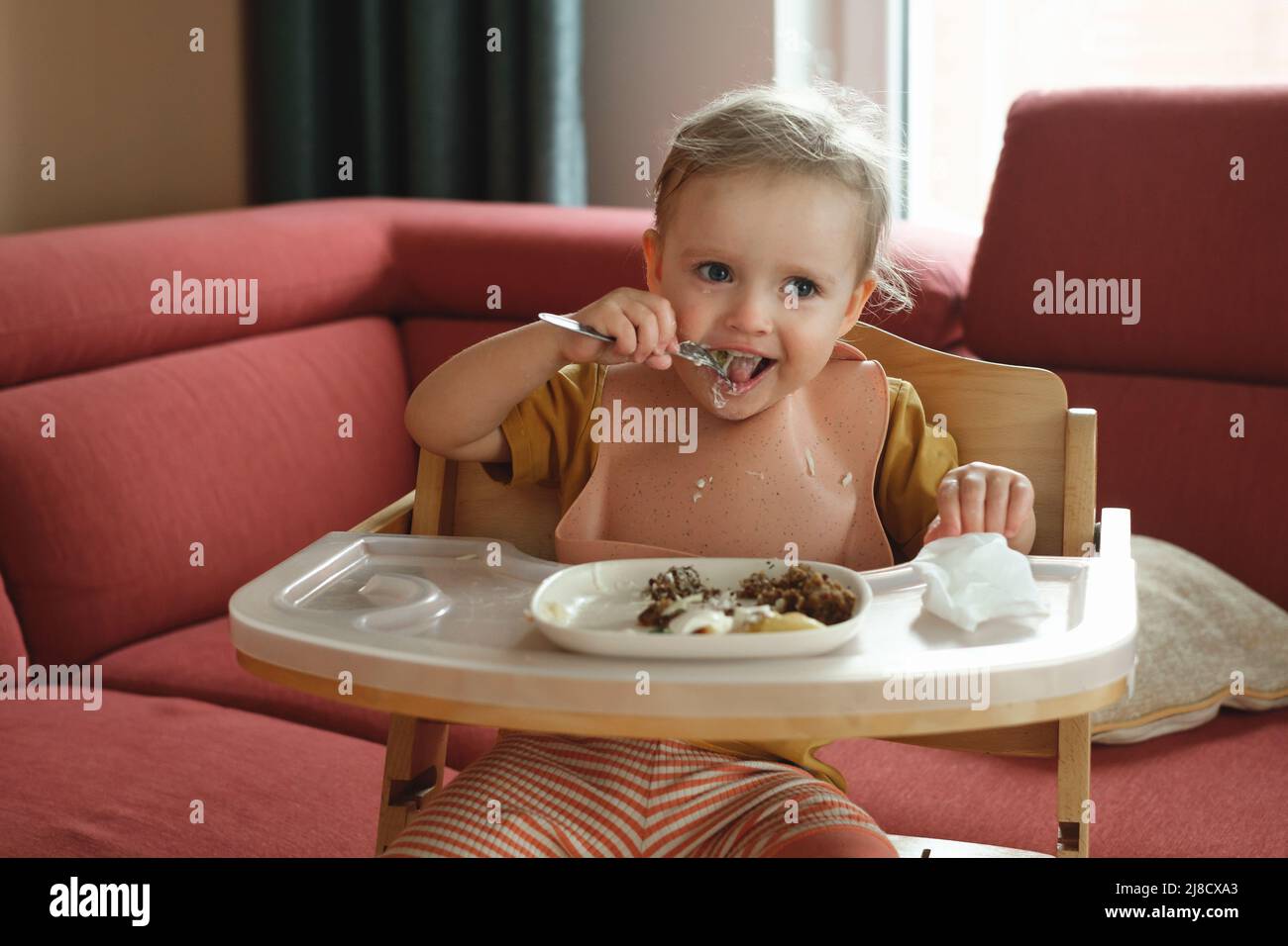 Toddler girl eating happily in high chair food by herself in the room ...