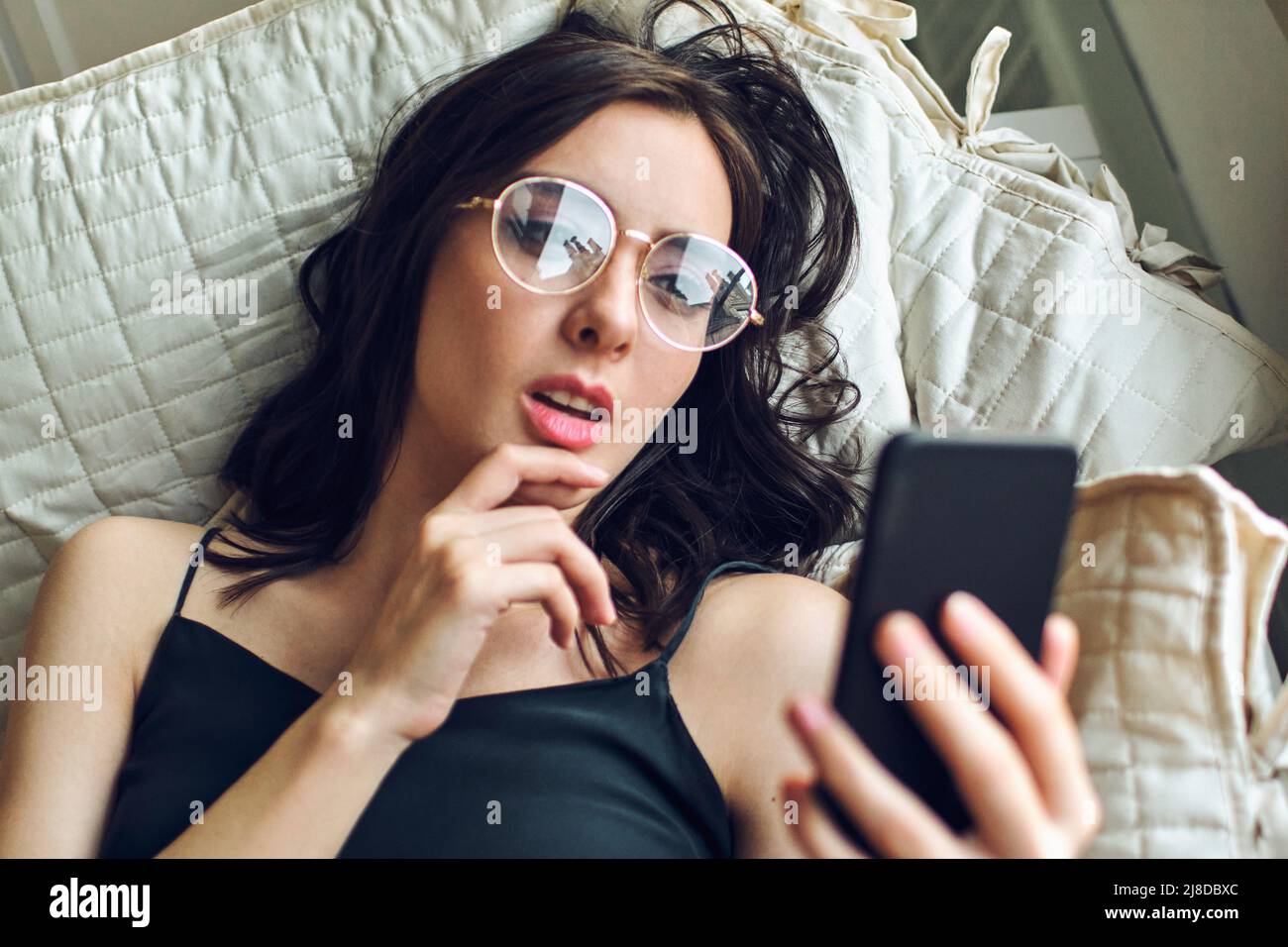 young adult woman hand on chin using mobile phone while lying in bed ...