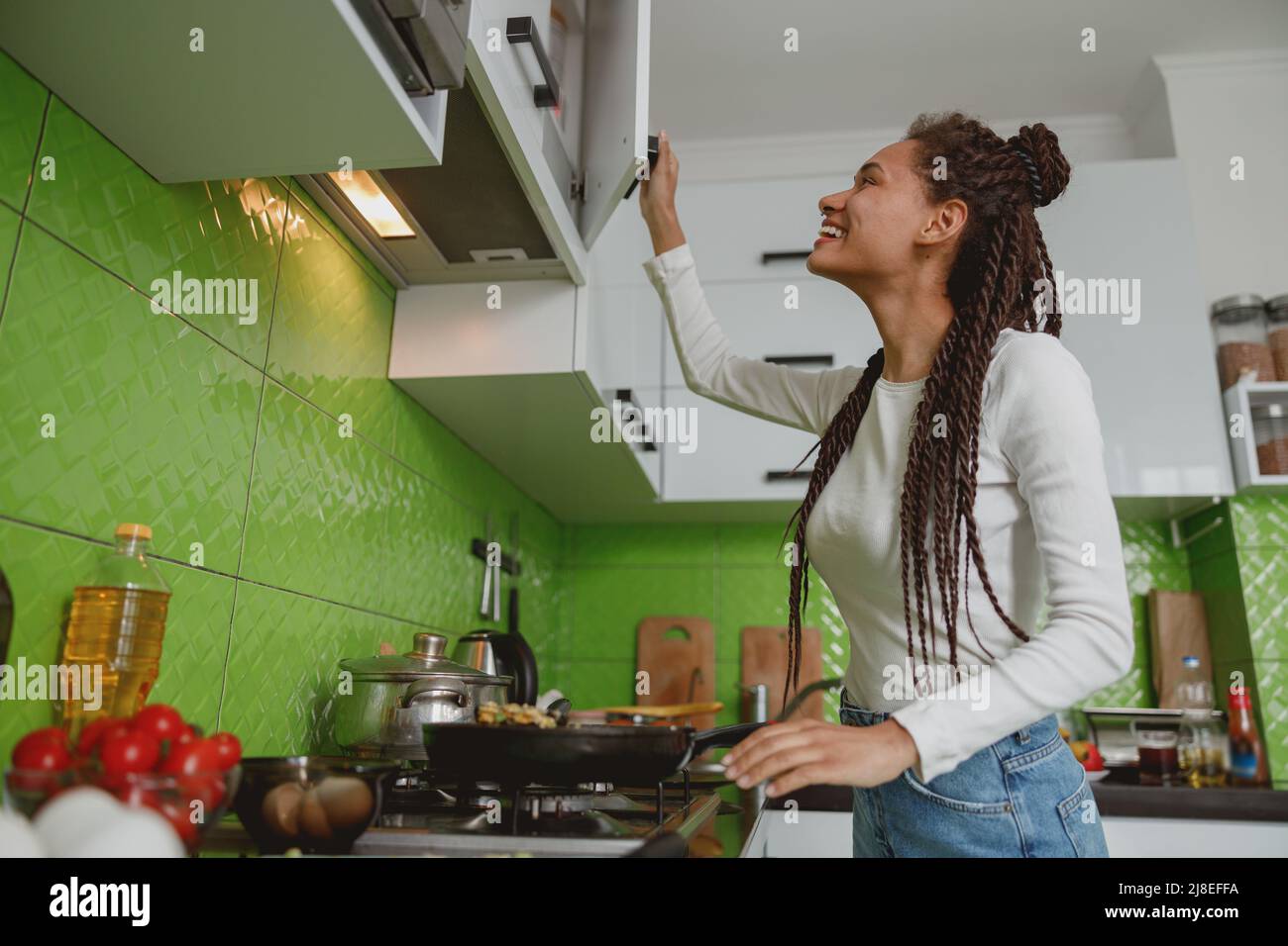 Smiling happy young female with braids opens kitchen cabinet, woman ...