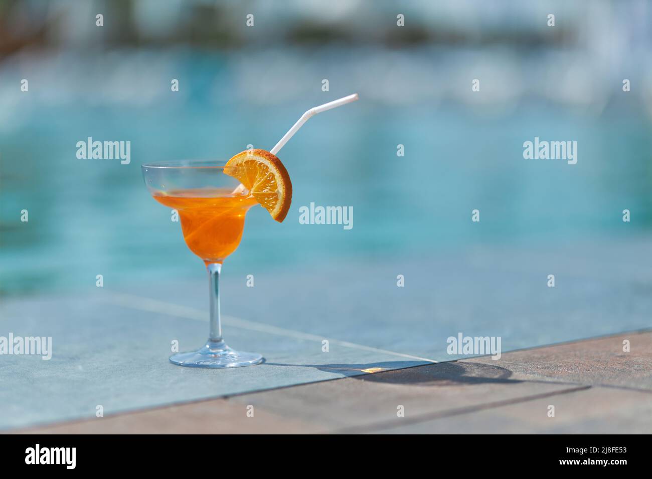 Tasty orange cocktail in glass located on grey tile edge of swimming pool in sunny afternoon ...