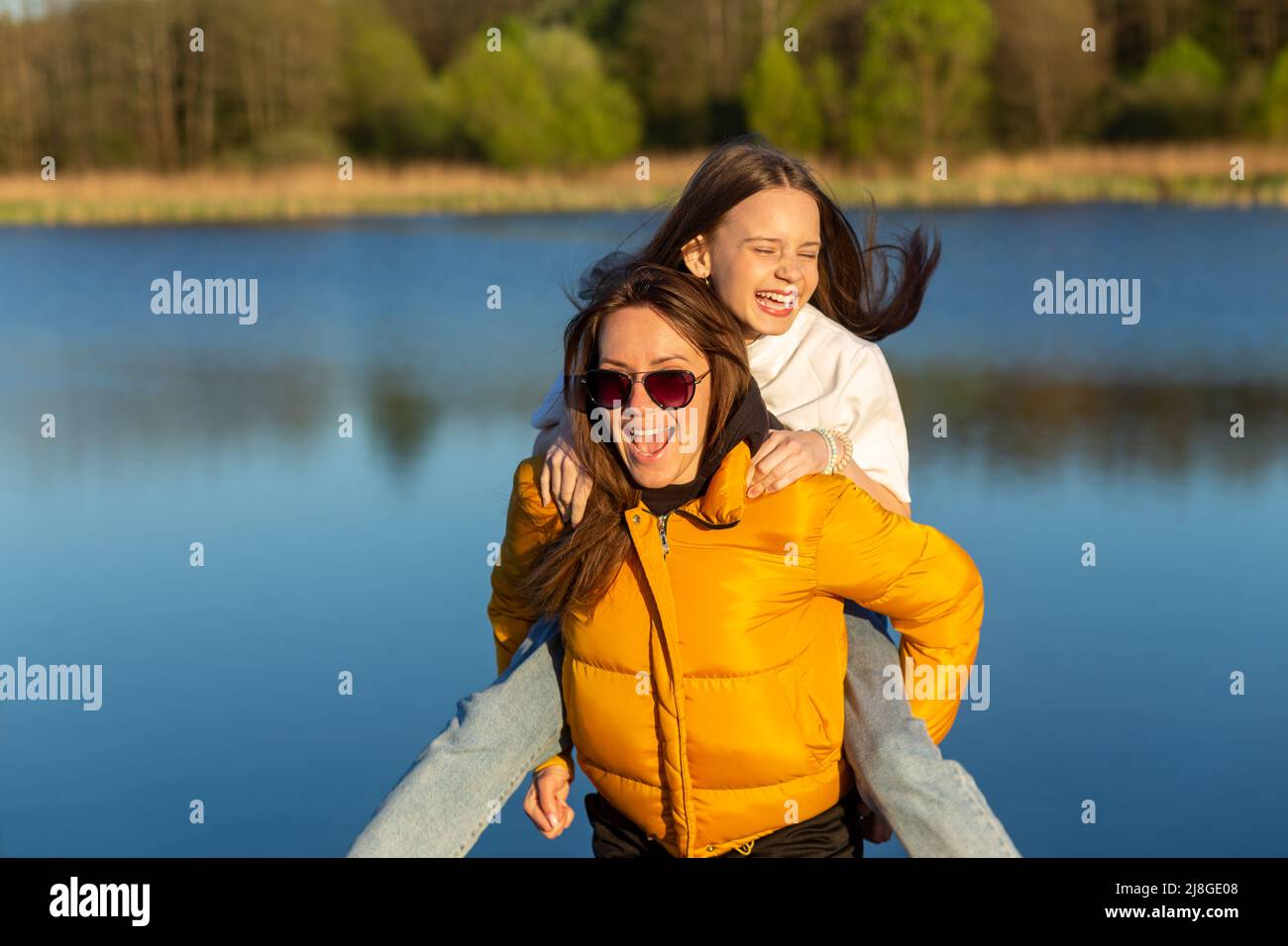 Playful mother giving daughter piggy back ride at spring lake shore ...